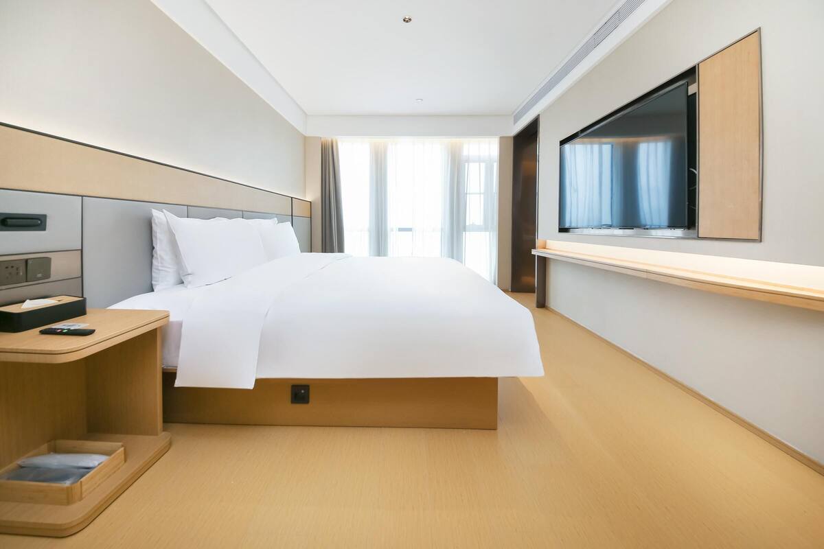 Quarto de hotel com cama, mesa de cabeceira, televisão e janela com cortinas.