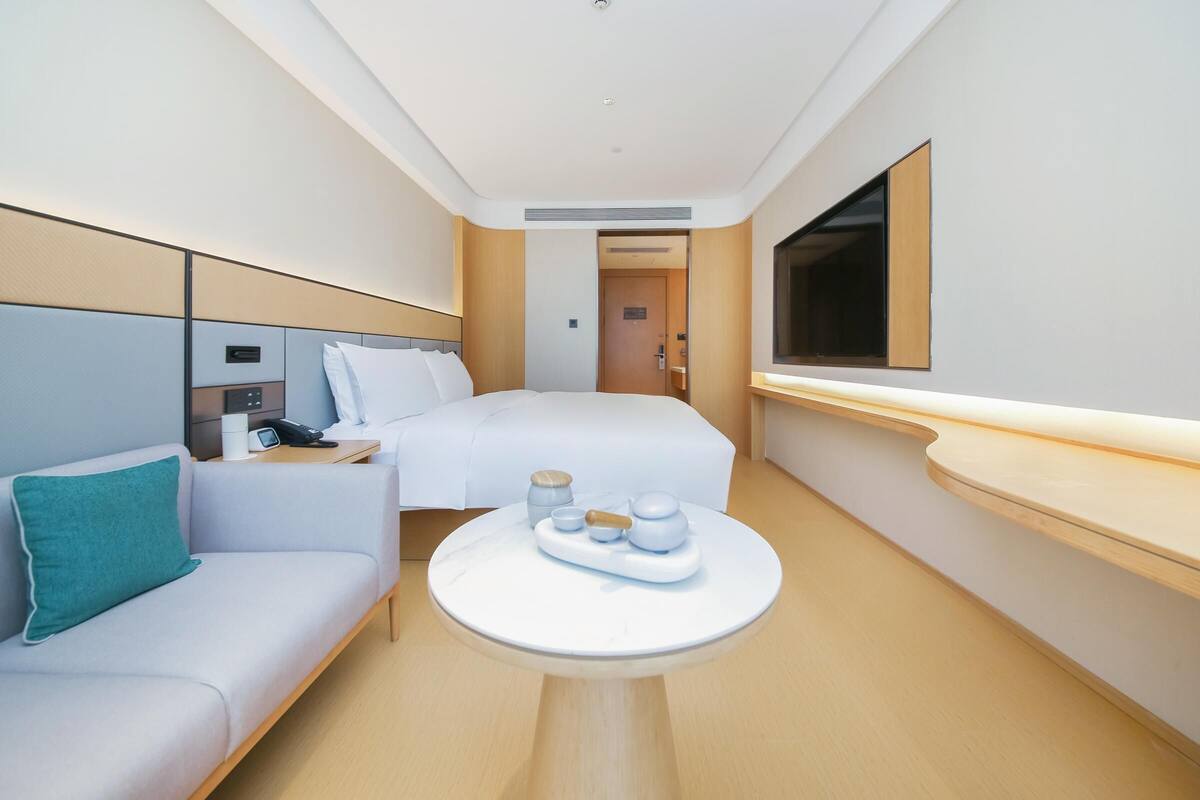 Quarto de hotel com uma cama, um sofá, uma mesinha com bandeja, uma televisão e uma porta que dá para outro quarto.