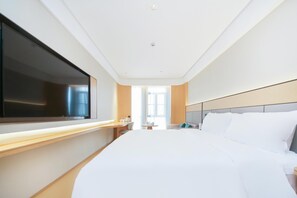 Quarto de hotel moderno com uma cama grande, televisão de tela plana na parede, mesa de cabeceira e vista para o corredor.