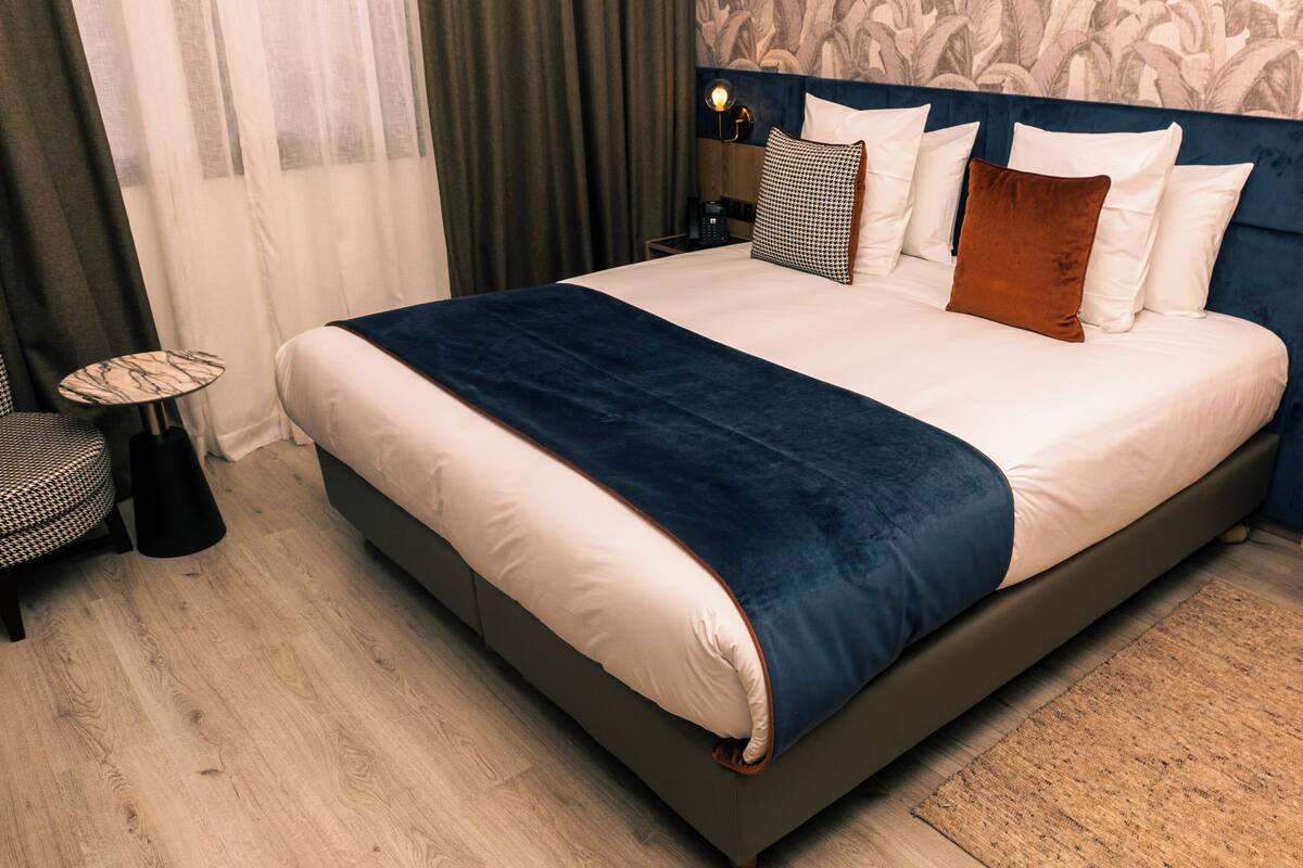 Uma cama grande com cabeceira escura, roupa de cama branca e várias almofadas. Há uma mesa de cabeceira com abajur, uma pequena mesa redonda e uma mesinha de cabeceira de madeira com gaveta.