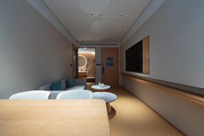 Quarto de hotel com chão de madeira, sofá, mesa redonda pequena, TV fixada na parede e porta para um banheiro.