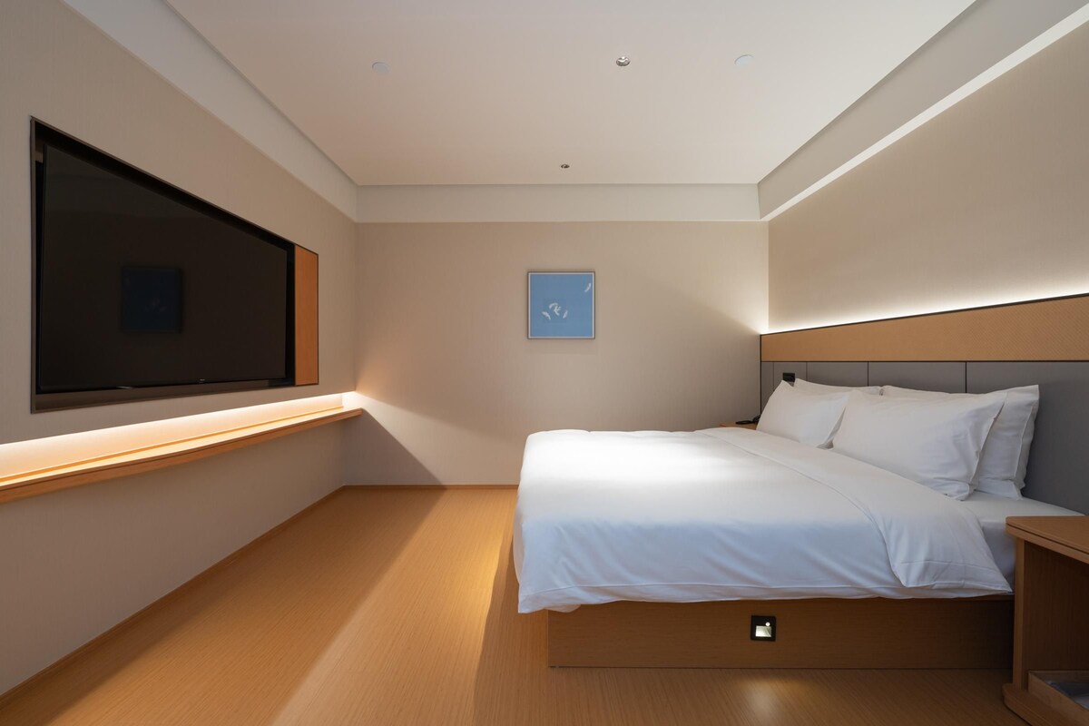 Quarto moderno com uma janela grande, uma cama, uma mesinha de cabeceira e uma TV fixada na parede.