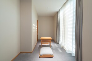 Um corredor de hotel estreito com um banco de madeira e uma pequena mesa, janelas amplas com cortinas finas e piso coberto de carpete.
