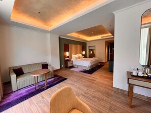 Quarto de hotel com cama, sofá, mesa redonda pequena, cadeira, espelho e piso de madeira.