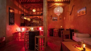 Zona de bar com banquetas, balcão de madeira e um grande barril. Há uma cama com cabeceira branca e uma mesa com uma vela.