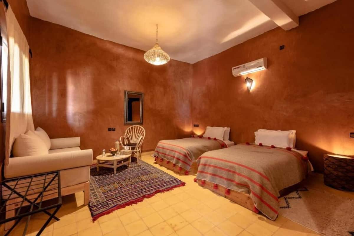 Quarto com duas camas, um sofá, uma mesa pequena, uma cadeira, luminária pendente e aparelho de ar condicionado.