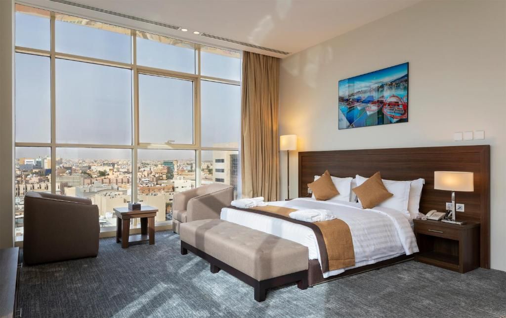 Quarto de hotel com uma janela grande, cama com roupa branca, cabeceira marrom, mesa de cabeceira com abajur e vista para a cidade.