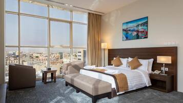 Quarto de hotel com uma janela grande, cama com roupa branca, cabeceira marrom, mesa de cabeceira com abajur e vista para a cidade.