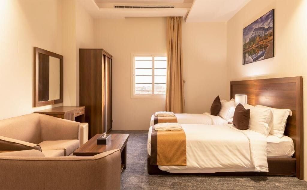 Quarto de hotel com duas camas, um sofá, uma escrivaninha, um espelho, uma janela com cortinas e um quadro na parede.