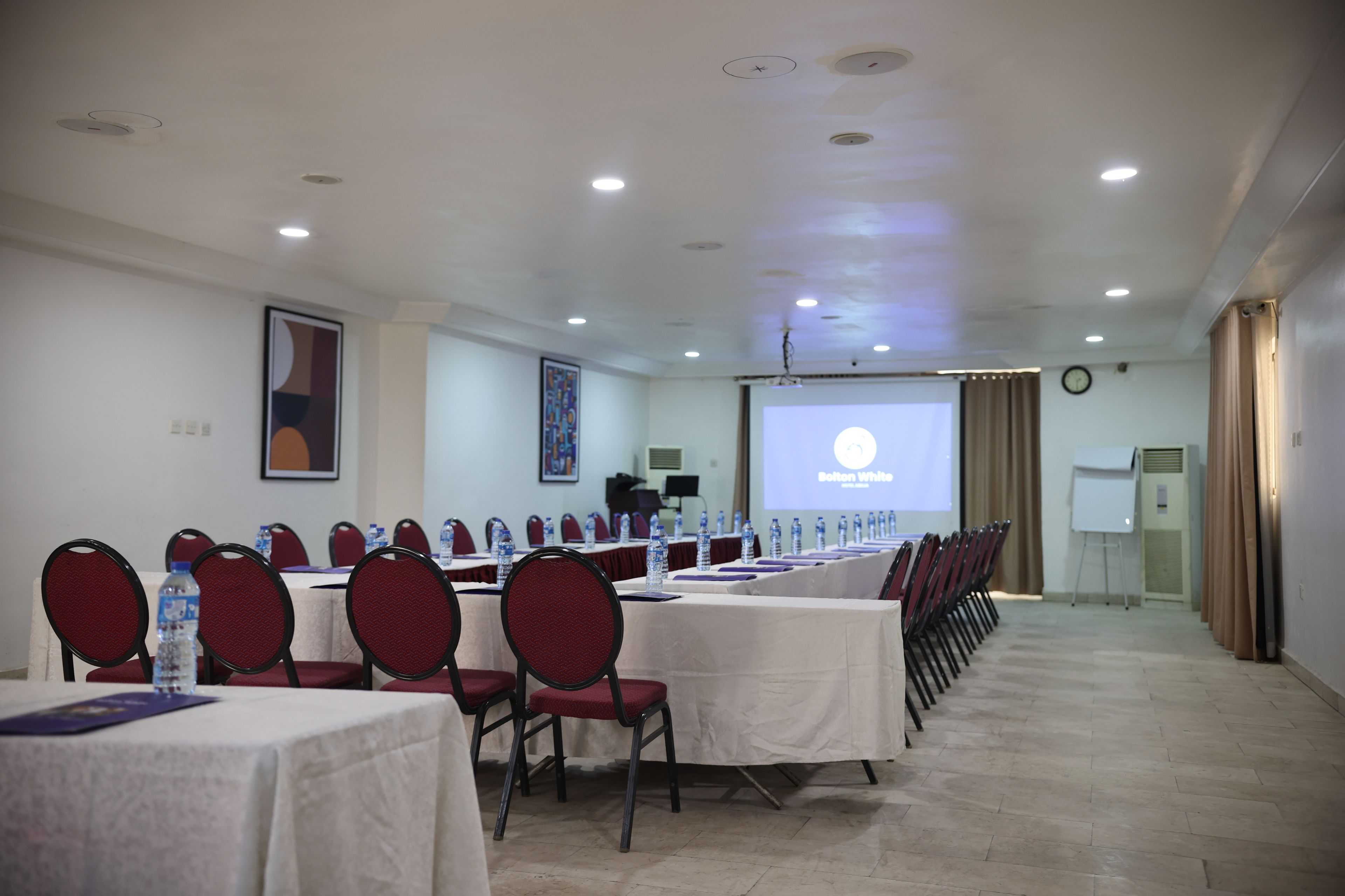 Uma sala de conferência com mesas, cadeiras, uma tela de projetor e uma tela para apresentação.