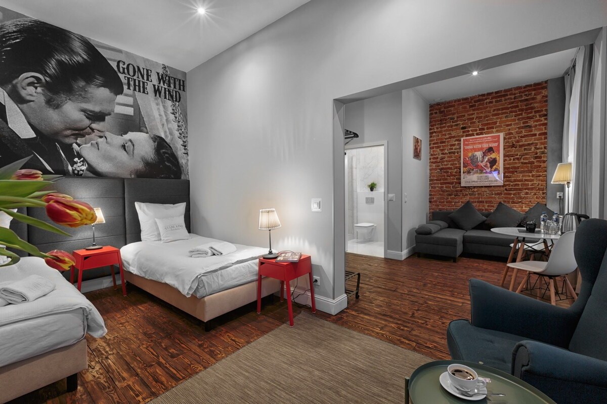 Quarto de hotel moderno com cama, mesa de cabeceira vermelha, mesa de centro, sofá e área de jantar.