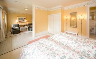 Quarto com cama, escrivaninha, duas cadeiras, guarda-roupa e banheiro visível por uma porta aberta.