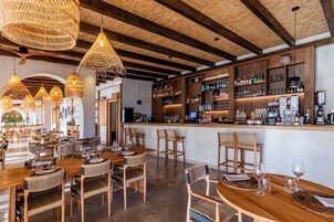 Um restaurante com mesas e cadeiras de madeira, um espaço de bar com prateleiras cheias de garrafas e luminárias de vime suspensas.
