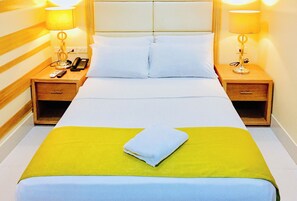Quarto de hotel com uma cama, duas mesas de cabeceira, um abajur em cada mesa, um controle remoto e um telefone em uma das mesas.