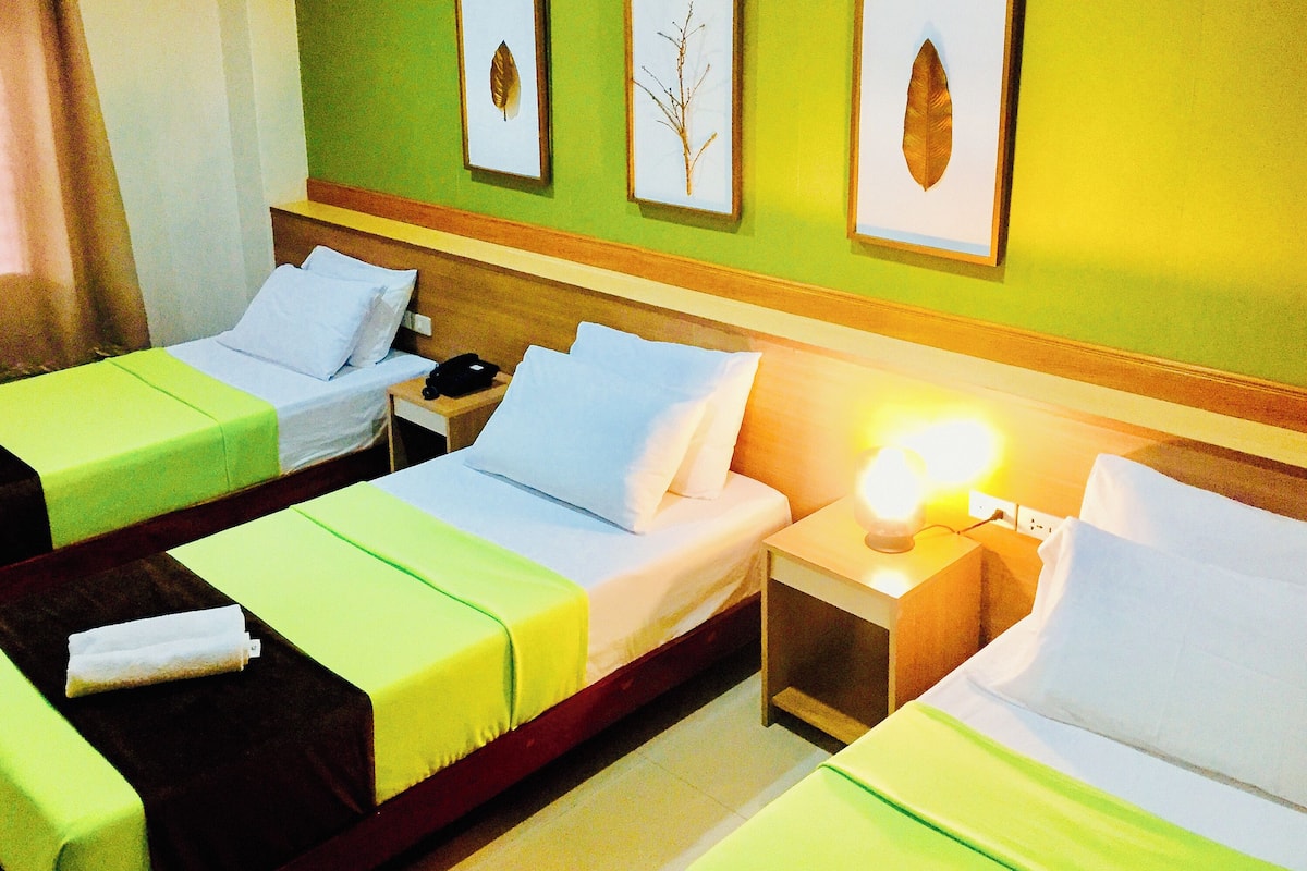 Quarto de hotel com duas camas, cada uma com colcha verde e branca, mesa de cabeceira com abajur e arte com folhagens emoldurada na parede.