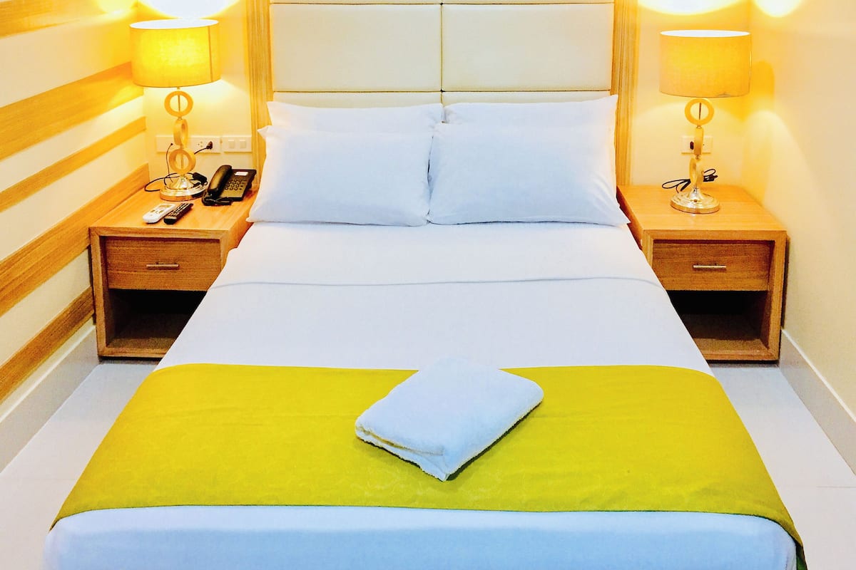 Quarto de hotel com uma cama, duas mesas de cabeceira, um abajur em cada mesa, um controle remoto e um telefone em uma das mesas.