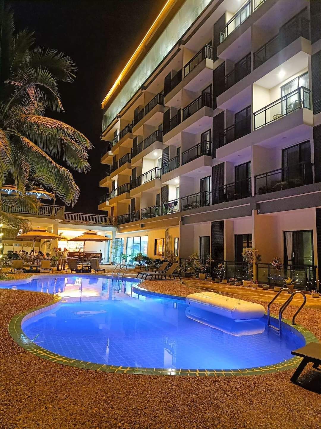 Um hotel com vários andares, piscina, área de estar externa e palmeiras.