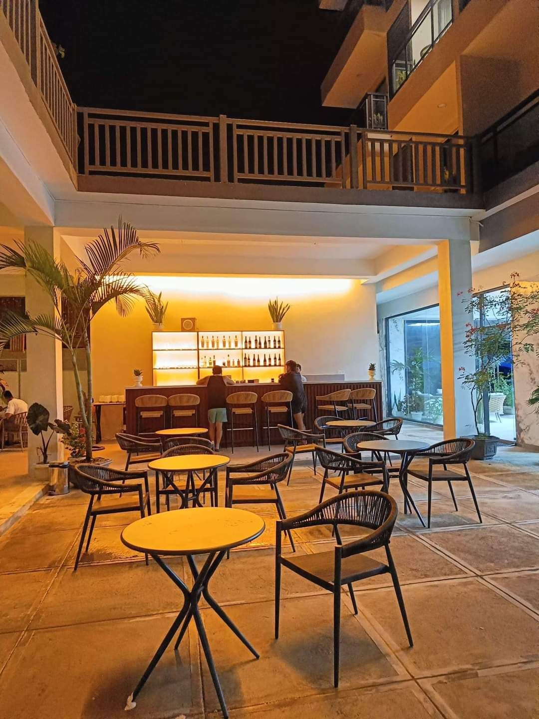 Uma área de estar externa com mesas e cadeiras, um bar com balcão, e uma varanda com vista para a área de estar.