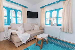Uma sala de estar com um sofá bege, um banquinho de madeira, um tapete azul estampado e uma TV fixada na parede.