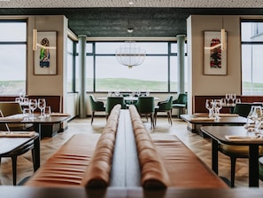 Restaurante com área de estar em um longo banco, mesas com cadeiras e grandes janelas com vista para o exterior.