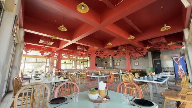 Um restaurante com mesas e cadeiras, vigas de teto vermelhas e luminárias pendentes.