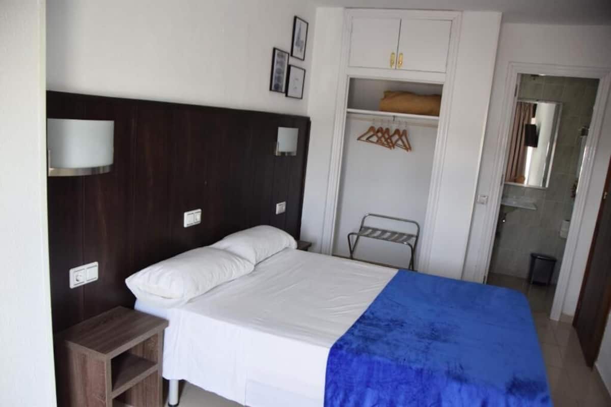 Uma cama de solteiro com colcha branca e azul, uma mesinha de cabeceira de madeira, um guarda-roupa com espaço para pendurar roupas e um espelho.