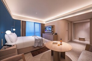Quarto de hotel moderno com uma cama grande, televisão de tela plana, mesa de centro redonda e área de estar com sofá e poltronas.