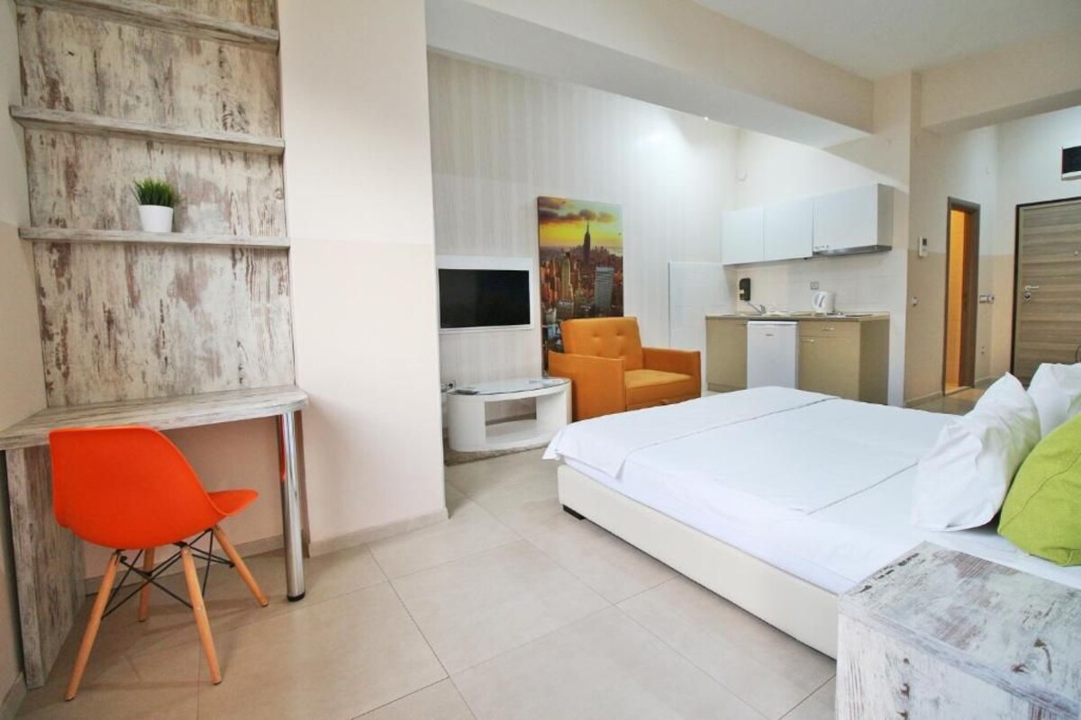 Quarto de hotel moderno com cama, escrivaninha, cadeira, televisão e cozinha compacta.