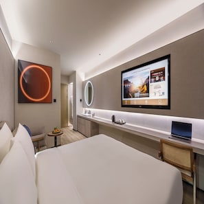 Quarto de hotel moderno com uma cama grande, uma escrivaninha com um laptop, uma TV fixada na parede e um candeeiro circular.