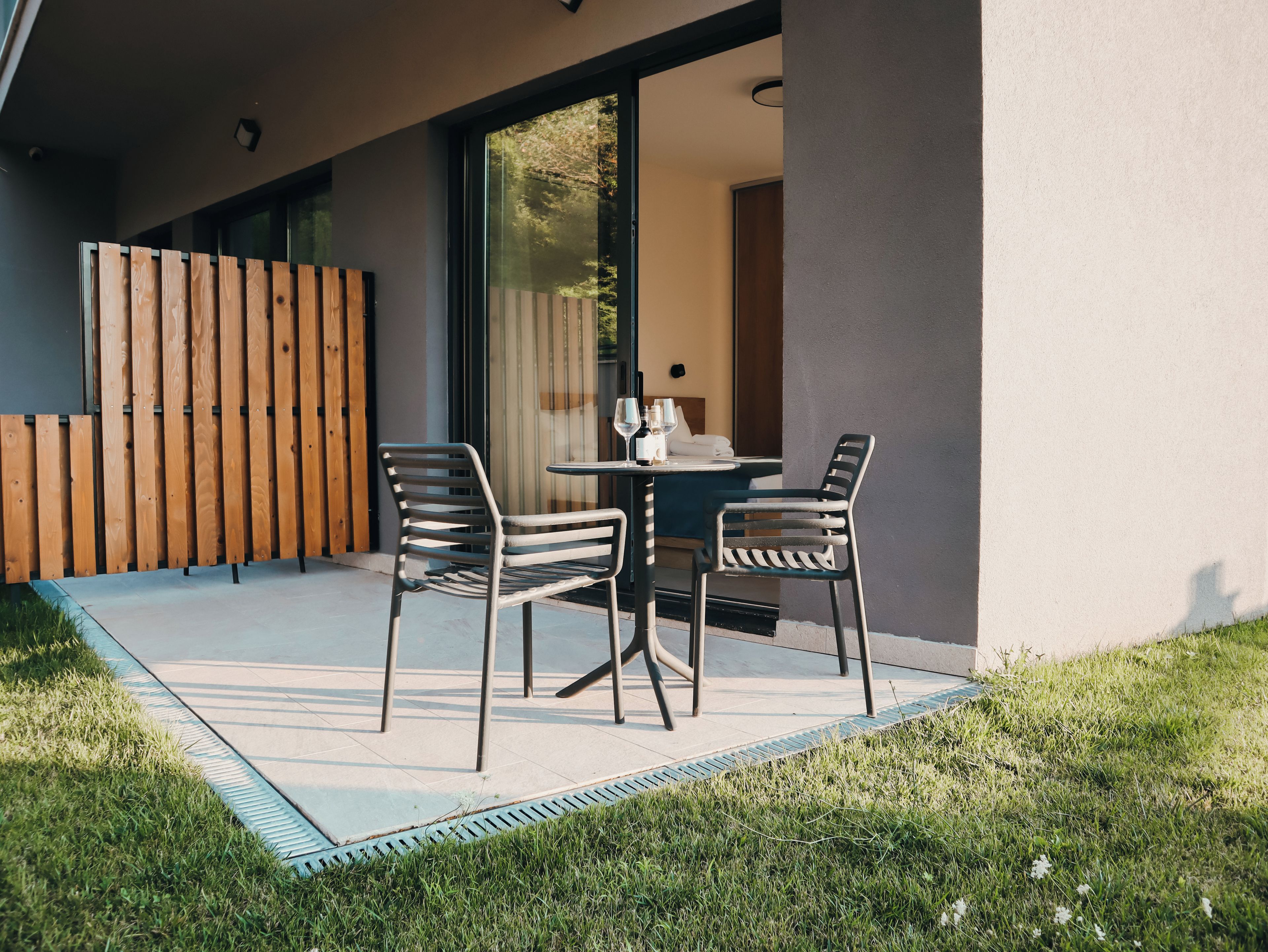 Um pátio externo com duas cadeiras, uma mesa, cerca de madeira e porta de vidro que dá para um espaço interno.