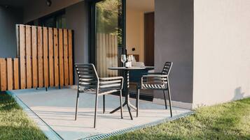 Um pátio externo com duas cadeiras, uma mesa, cerca de madeira e porta de vidro que dá para um espaço interno.