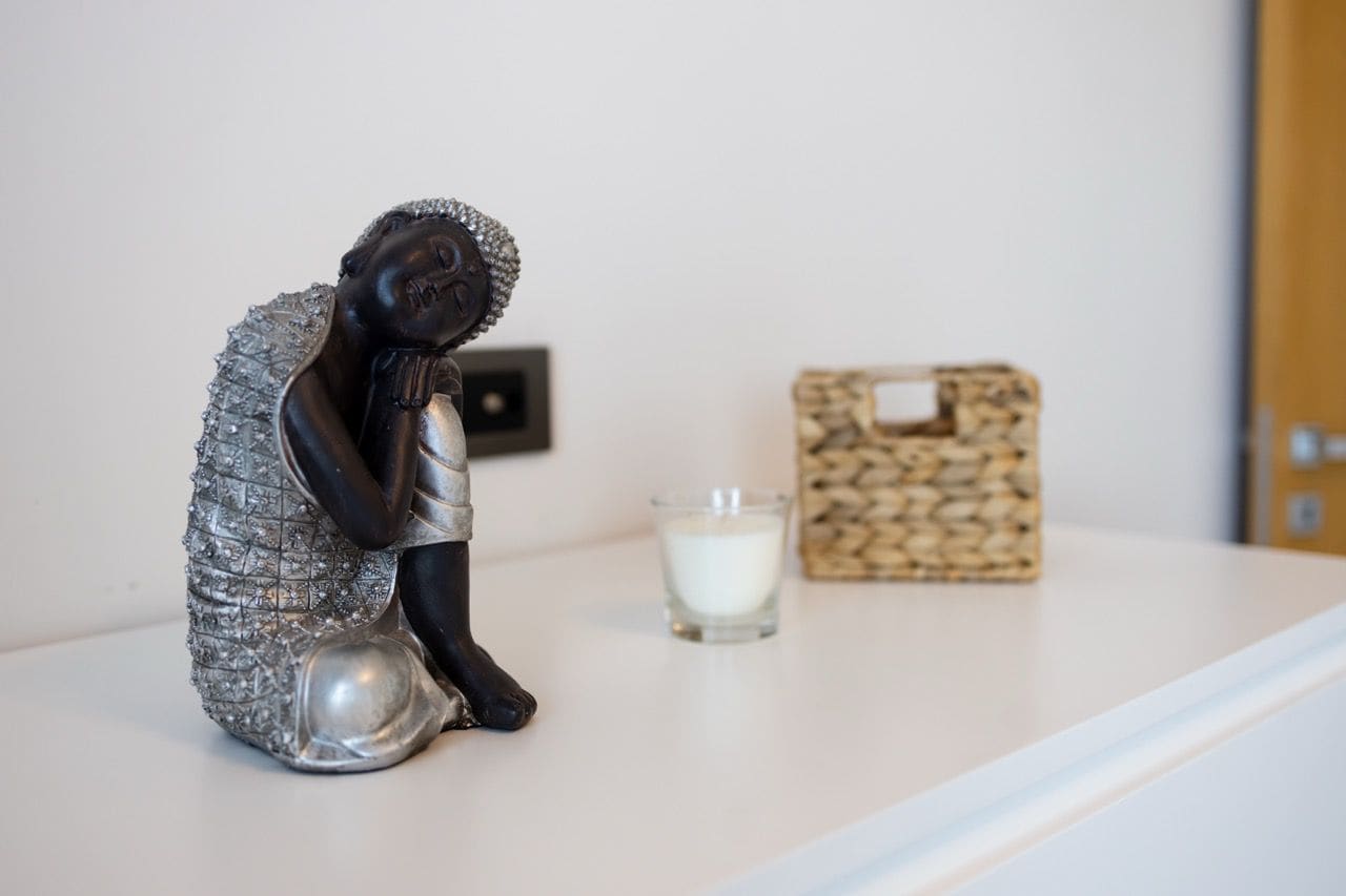 Uma estátua de uma figura contemplativa sobre uma superfície branca, uma vela e uma cesta de palha.