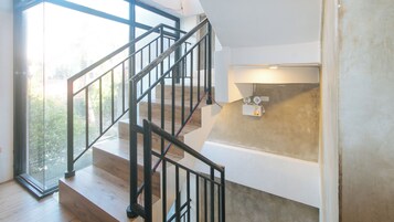 Escada com grades pretas, degraus de madeira e parede de concreto com luminária.