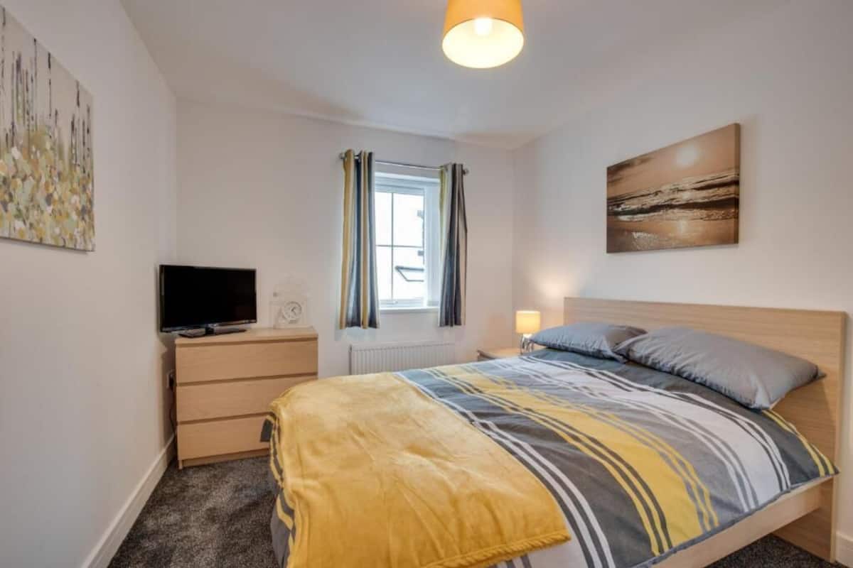 Quarto com cama, televisão, mesa de cabeceira, janela com cortinas e obras de arte nas paredes.