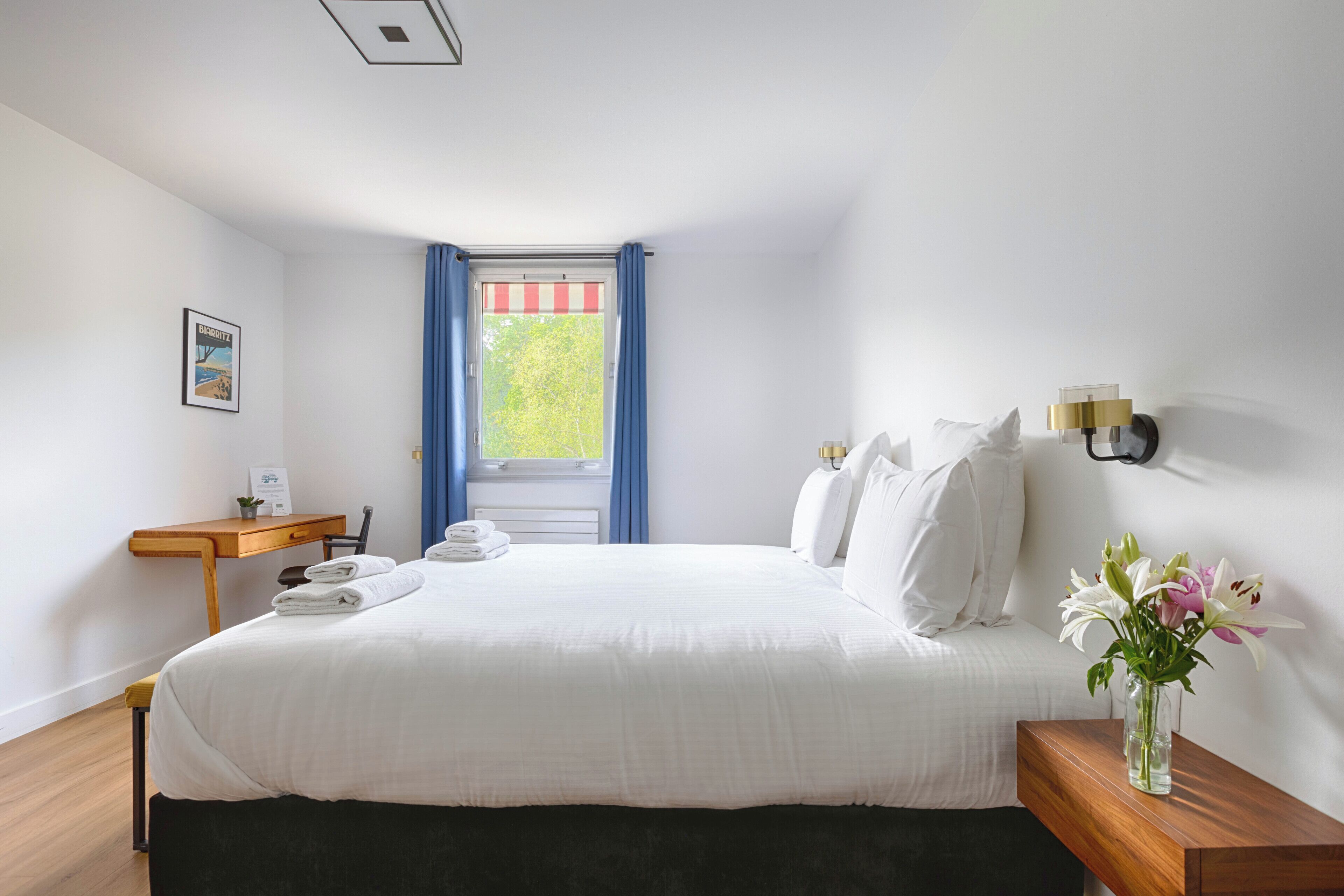 Quarto de hotel com cama, escrivaninha, cadeira, mesa de cabeceira, um vaso com flores e janela com cortinas.
