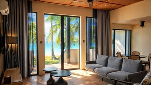 Uma sala de estar moderna com um sofá cinzento, uma mesinha redonda e uma poltrona verde. Há uma grande porta de vidro deslizante que dá para uma varanda com vista para palmeiras e o oceano.
