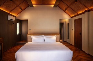 Quarto de hotel com cama grande, piso de madeira, teto inclinado com iluminação embutida, janela e porta.