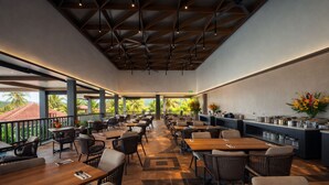 Uma ampla área de refeições ao ar livre com várias mesas e cadeiras, um balcão de bar e vista para palmeiras e um edifício com telhado vermelho.
