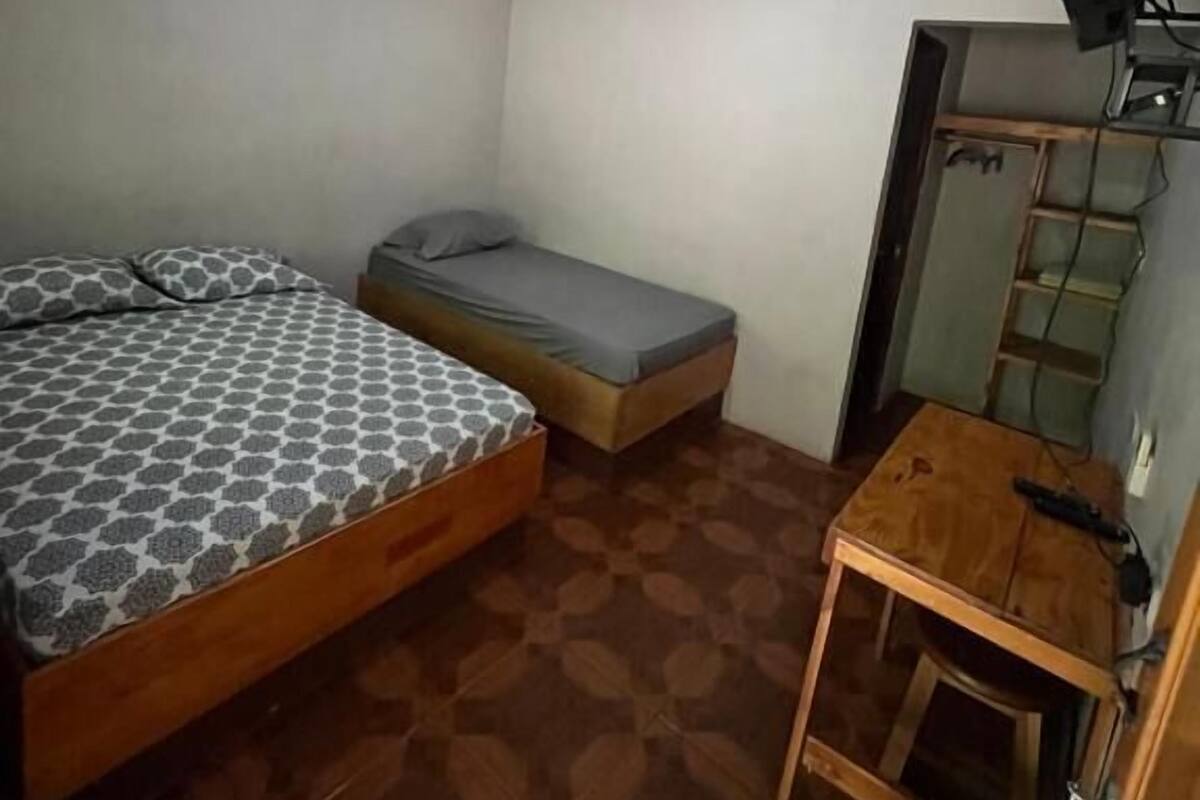 Quarto com uma cama, uma cama menor, uma mesa de madeira, uma TV e um ventilador de teto.