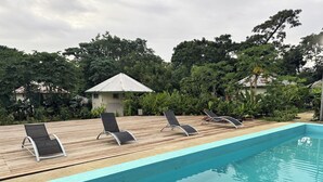 Piscina com quatro espreguiçadeiras, rodeada por vegetação exuberante e um pequeno edifício com telhado inclinado.
