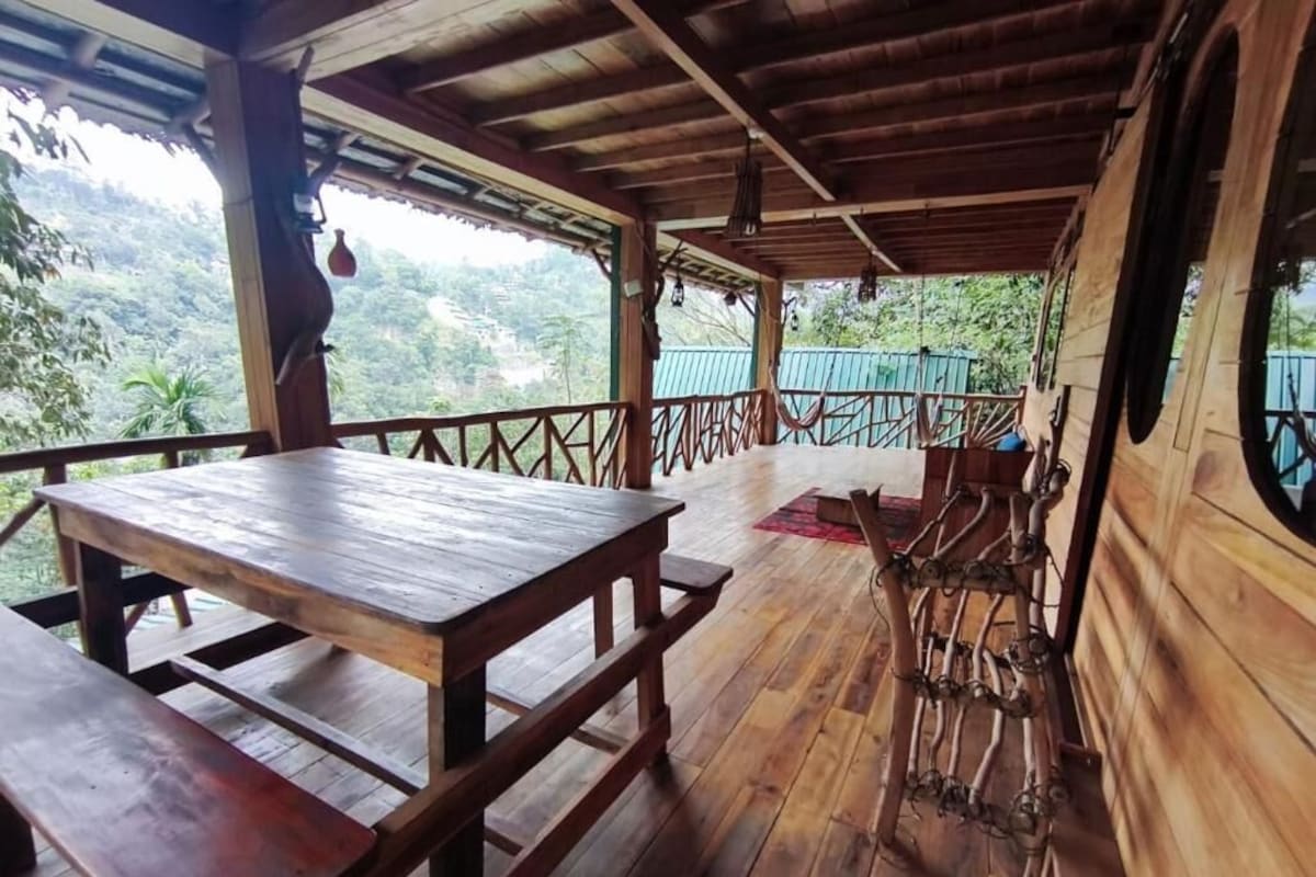 Um deck de madeira com uma mesa e bancos, cercado por um corrimão e vista para o verde.