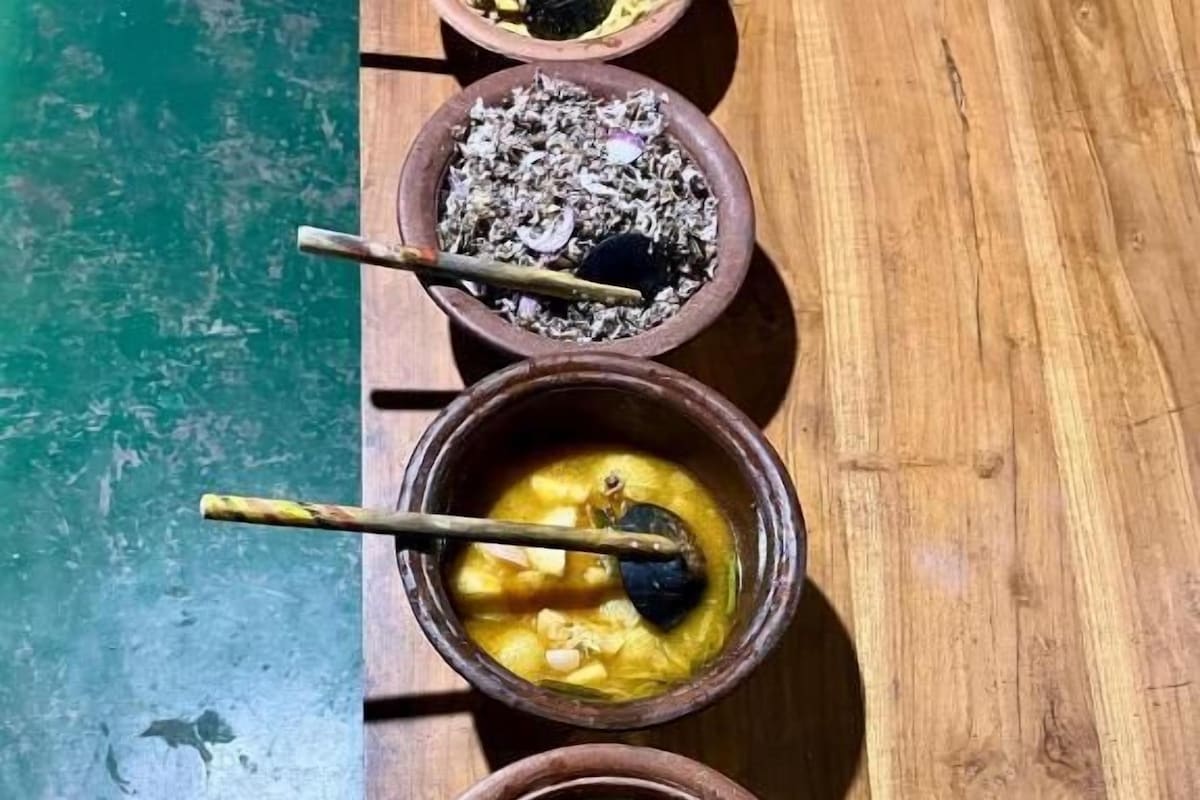 Uma mesa de madeira com quatro vasos de barro contendo diferentes alimentos, uma pessoa em pé com uma bengala, e um chão verde.