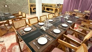 Uma sala de jantar com uma mesa comprida posta para várias pessoas, cadeiras de madeira e uma parede de destaque em pedra.