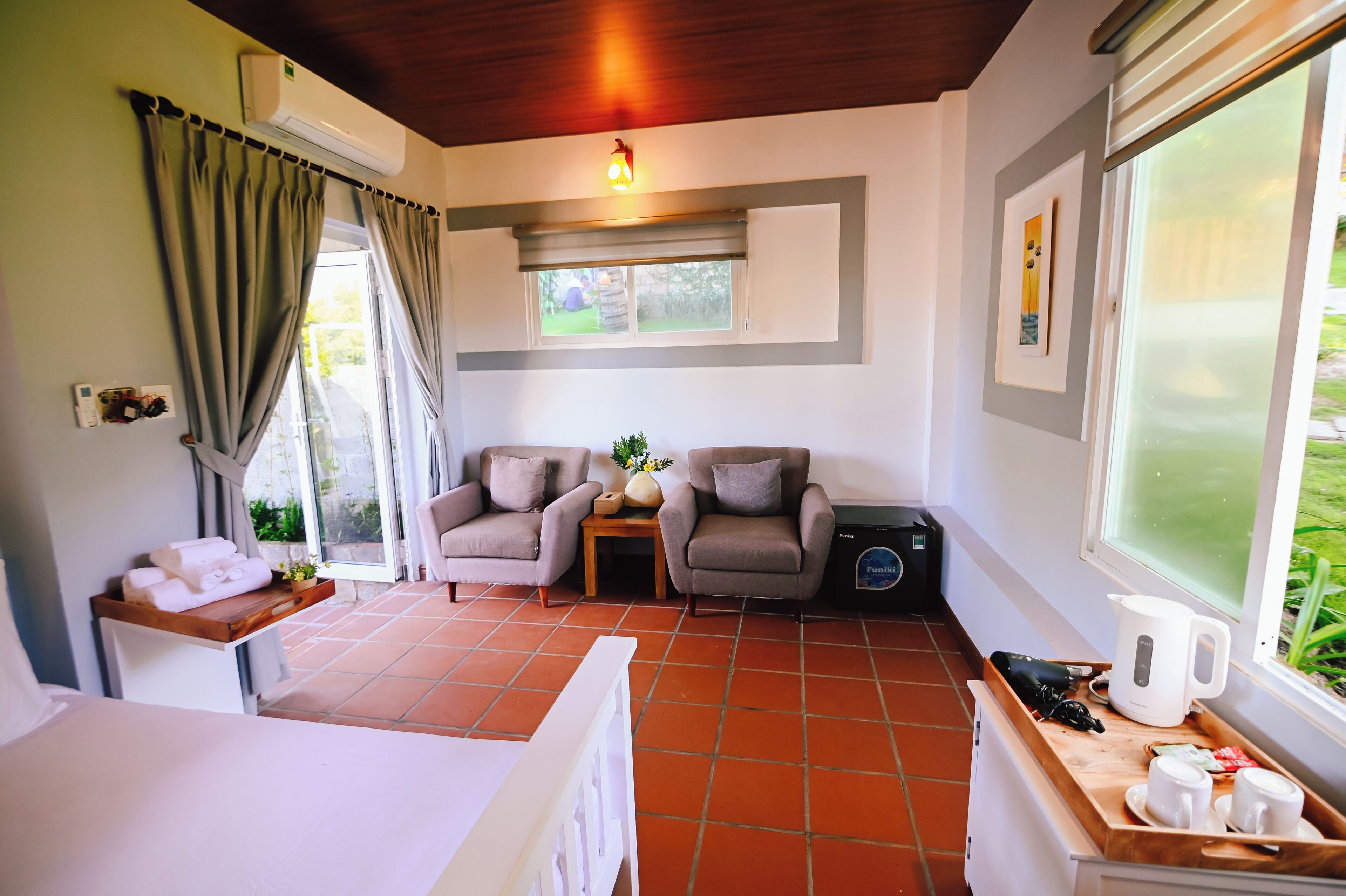 Um quarto com dois sofás, uma mesinha com uma planta, uma janela com cortinas, um radiador e um tabuleiro com chávenas e uma chaleira.