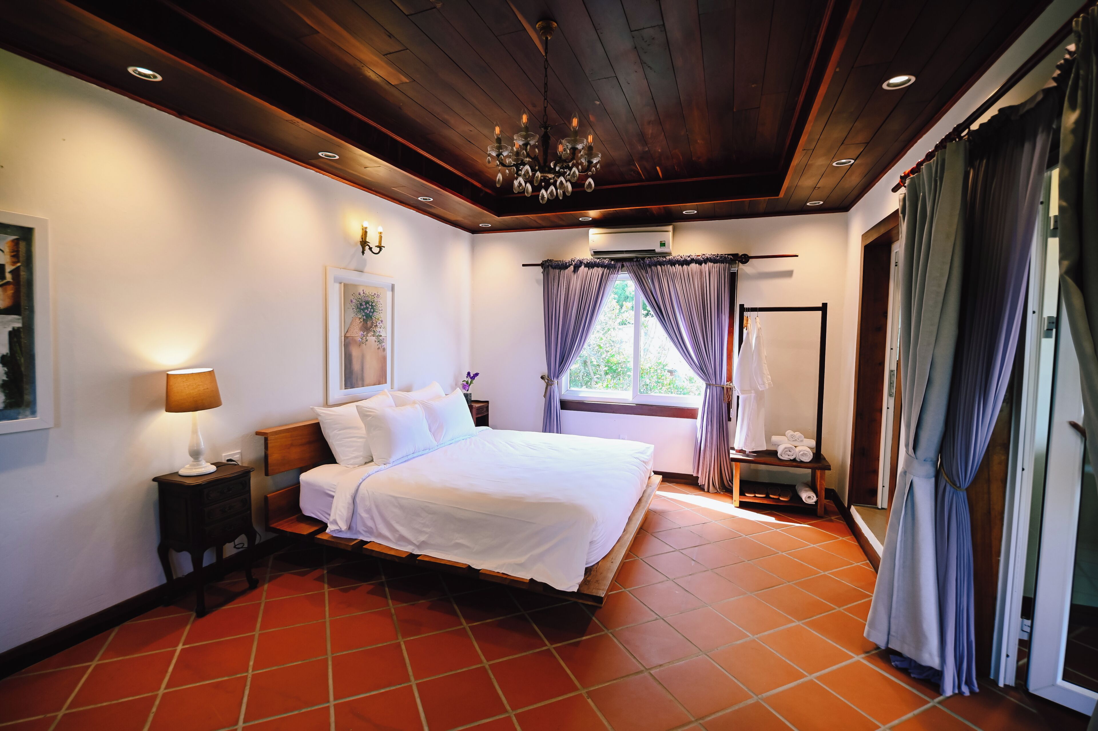 Quarto com teto de madeira, cama, mesa de cabeceira, abajur, janela com cortinas e porta.