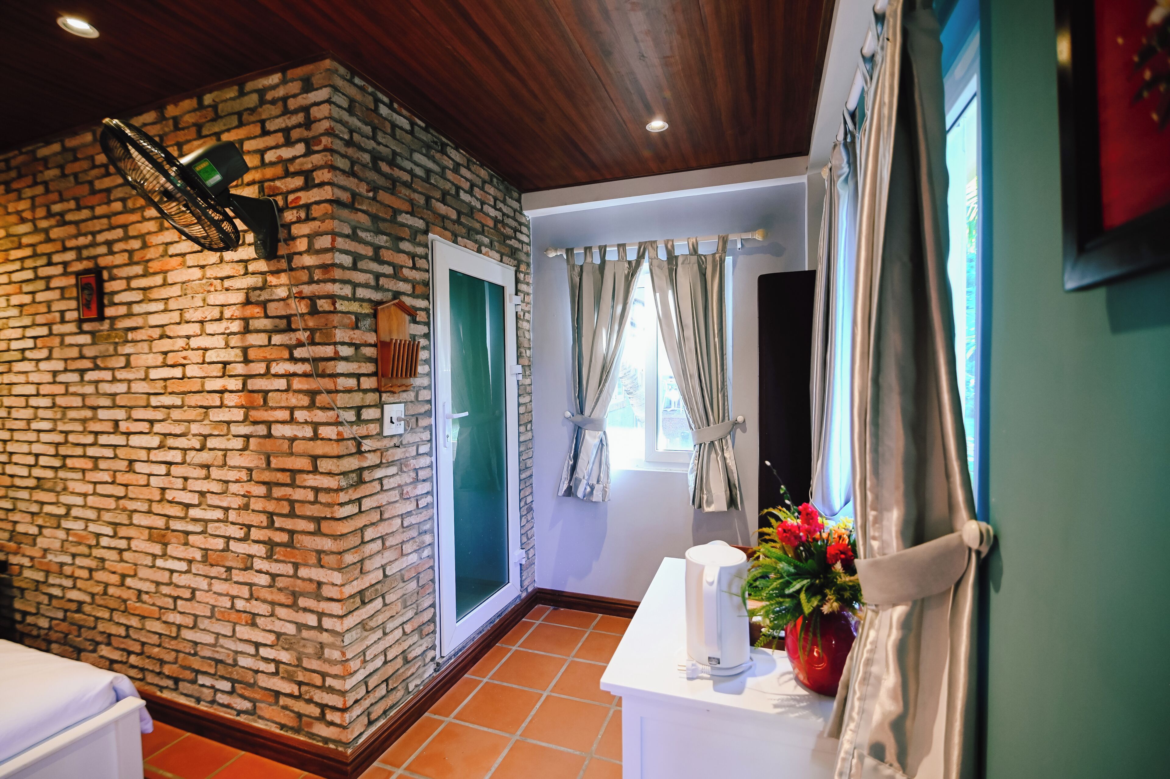 Quarto com parede de tijolos, radiador branco, janela com cortinas e um vaso com flores.