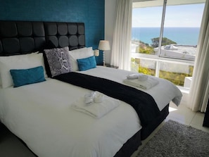 Uma cama grande com lençóis brancos, cabeceira preta e almofadas azuis. Há uma janela com vista para o oceano e edifícios.