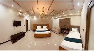 Quarto de hotel com uma cama grande, uma cama menor, uma escrivaninha, uma cadeira, uma TV e um ventilador de teto.