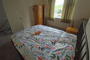 Quarto com colcha florida, guarda-roupa de madeira, radiador e janela com cortinas.