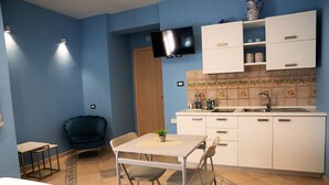 Quarto compacto com cama, mesa de jantar com cadeiras, pequena cozinha com pia e televisão fixada na parede.